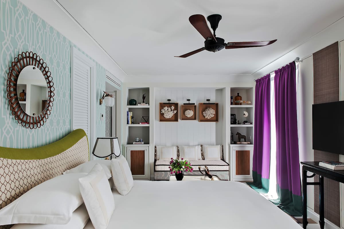 Quarto de hotel com cama, ventilador de teto, televisão, escrivaninha e janela com cortinas roxas.
