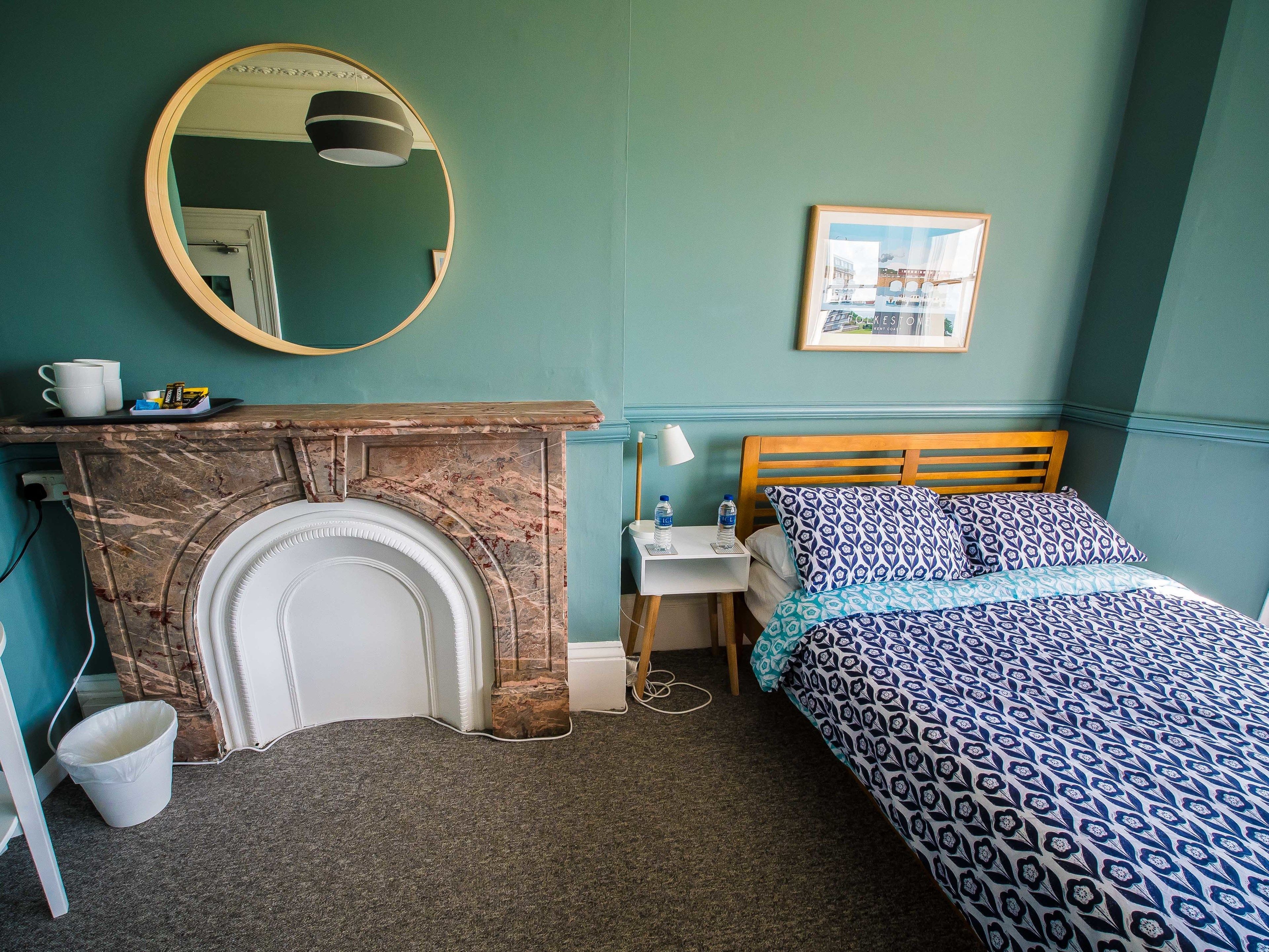 Quarto com lareira de mármore, cama com roupa de cama estampada, mesa de cabeceira com abajur e um quadro na parede.