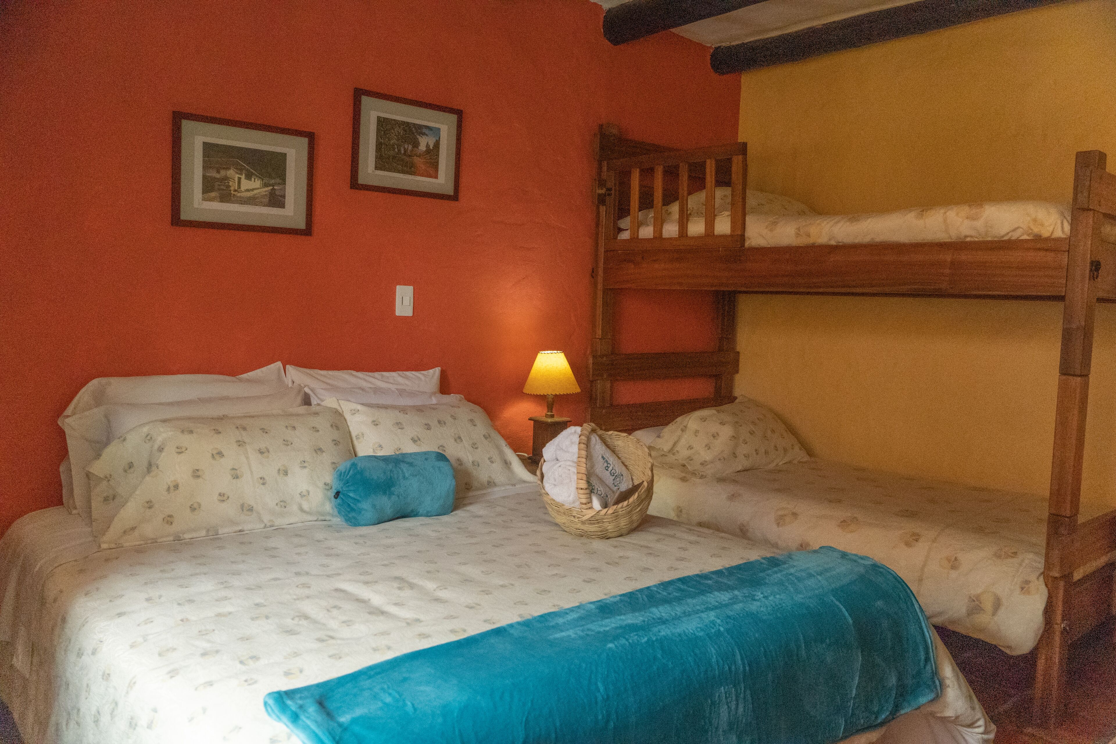 Quarto com duas camas, uma delas um beliche. Há uma luminária em um criado-mudo ao lado das camas, e uma cesta na cama de baixo.