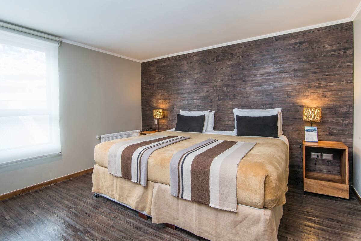 Quarto de hotel moderno com uma cama grande, cabeceira de madeira e duas luminárias laterais.