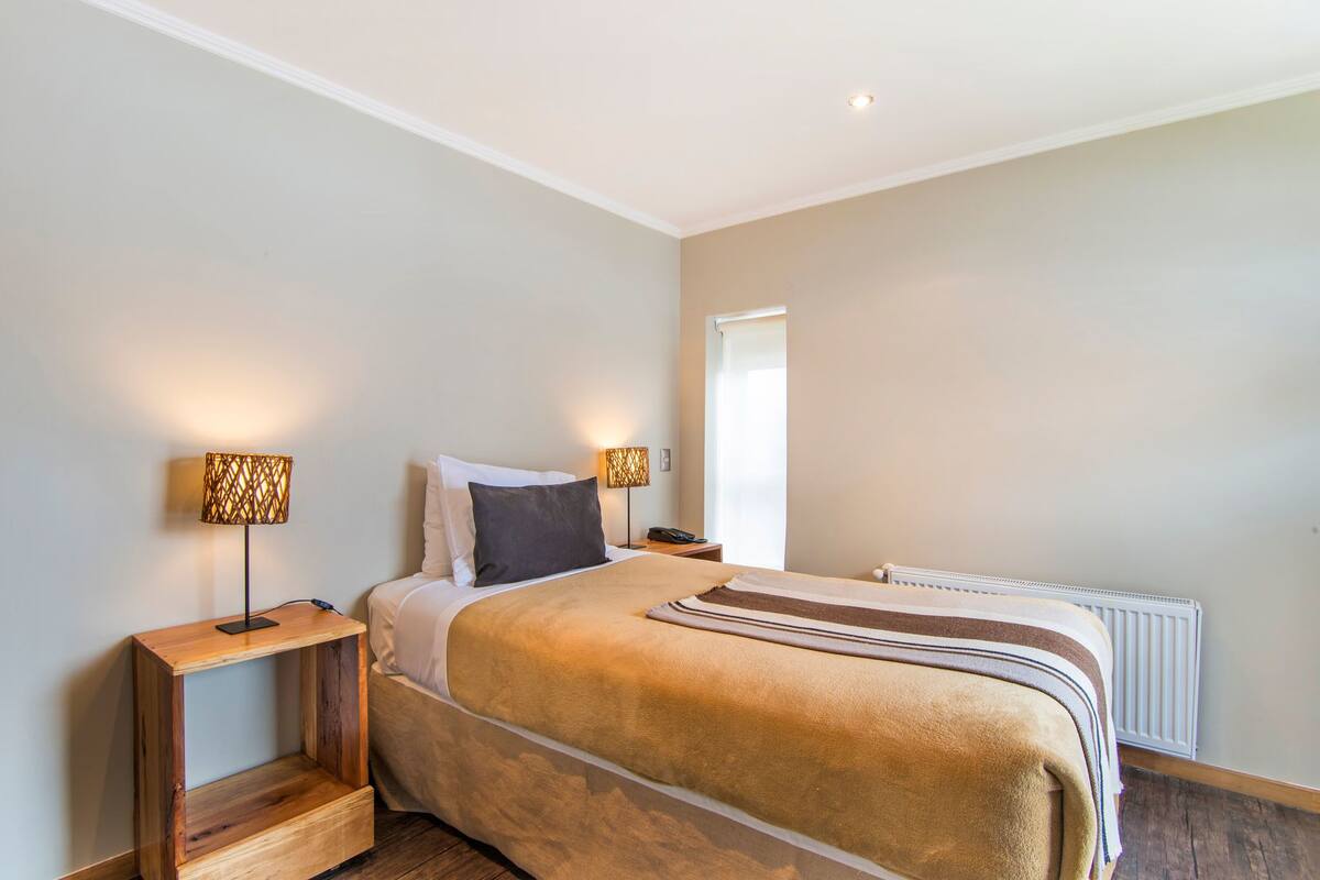 Uma cama bem arrumada com um cobertor bege e almofadas, uma mesinha de cabeceira de madeira com uma luminária e um radiador na parede.