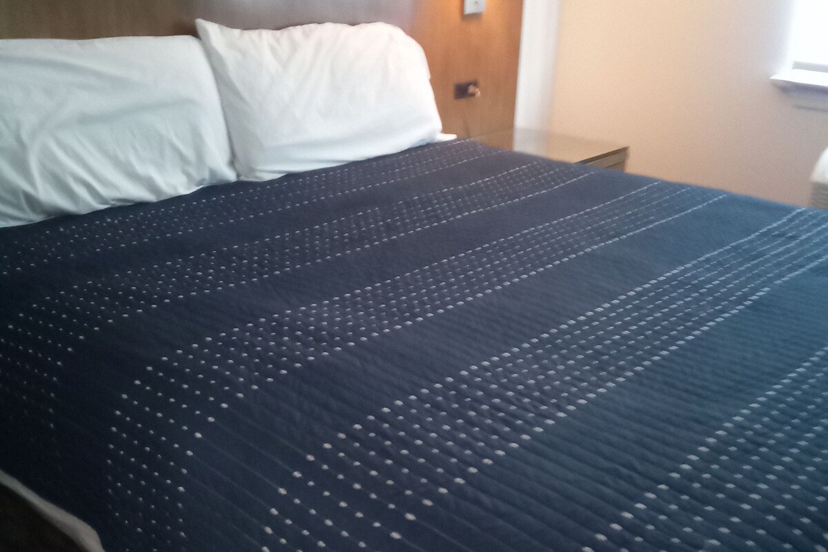 Cama com colcha escura e travesseiros brancos, cabeceira de madeira e luminária fixada na parede.