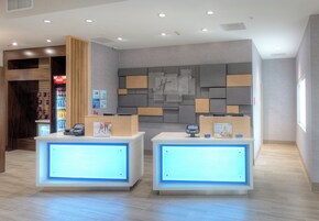 Uma receção de hotel com um ecrã digital, um refrigerador de Coca-Cola e uma parede com design geométrico.