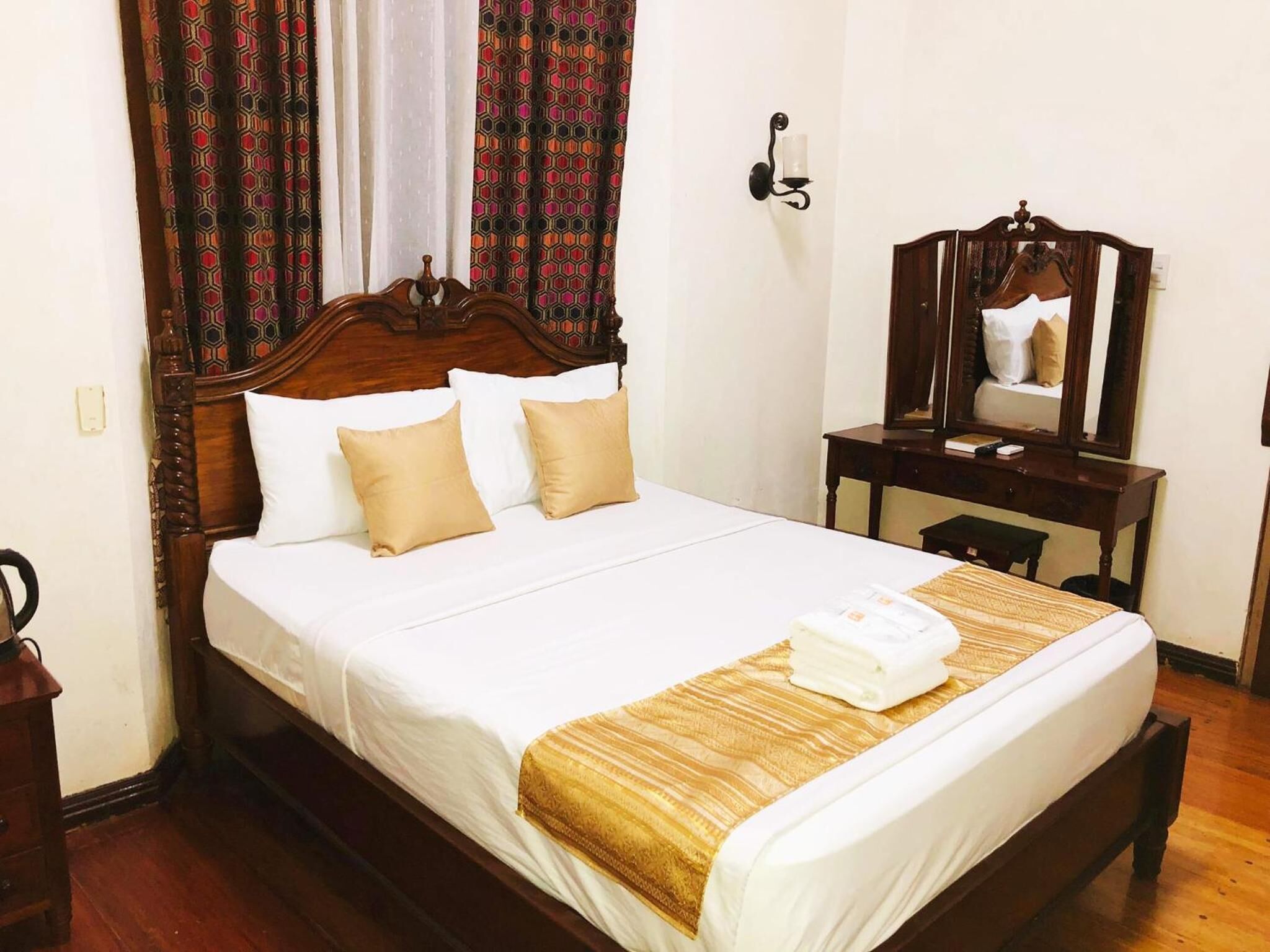 Uma cama bem arrumada com cabeceira de madeira, uma mesa de cabeceira com espelho e cortinas estampadas.
