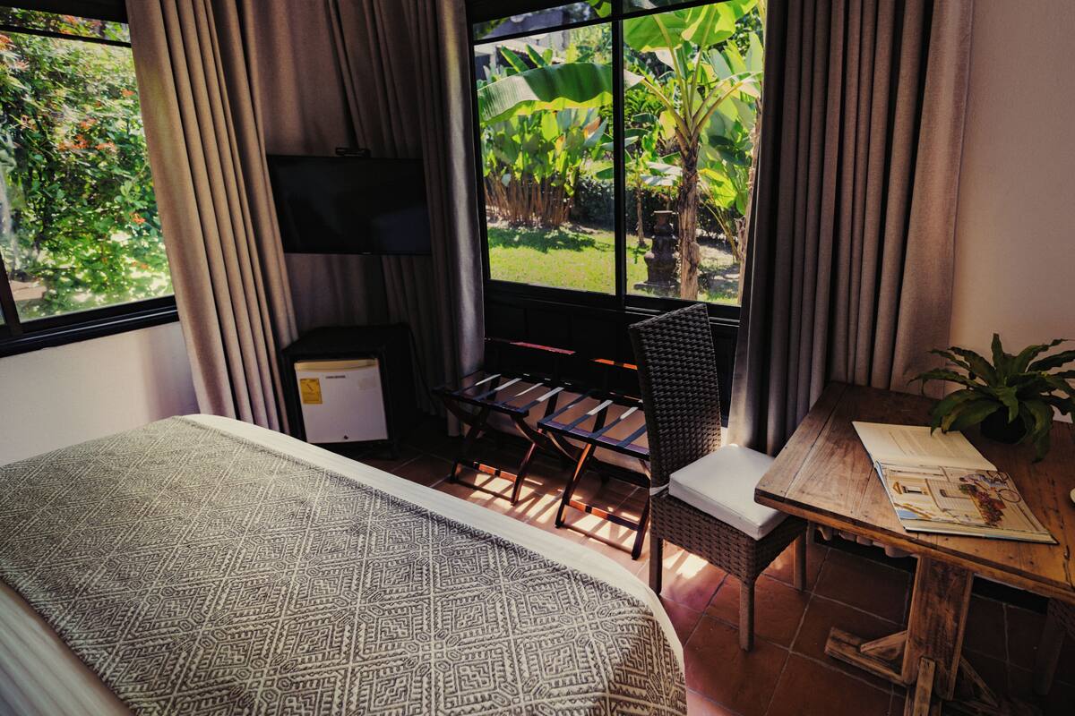 Quarto com cama, escrivaninha, cadeira, televisão e janela com cortinas.