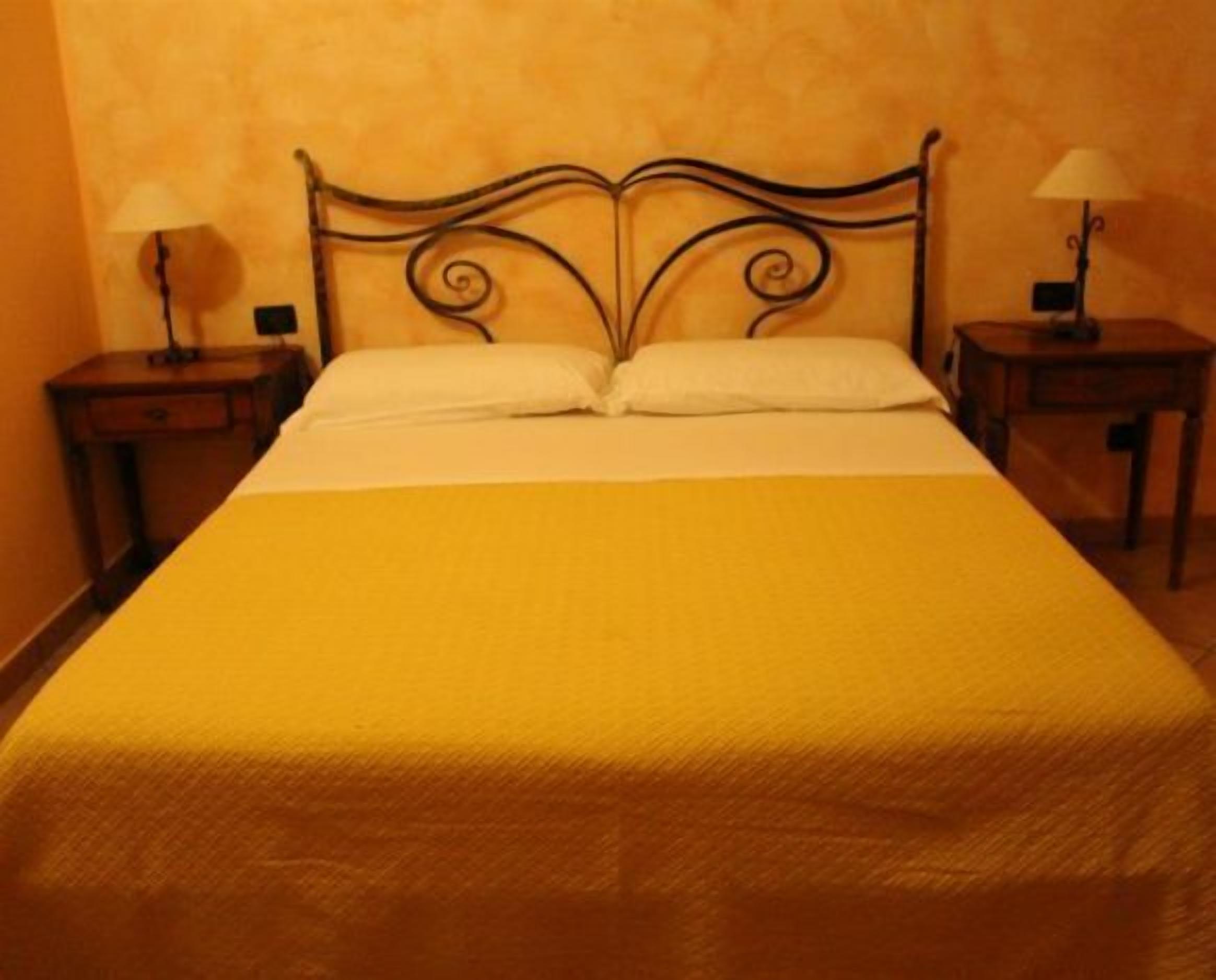 Uma cama com colcha amarela, cabeceira de metal e duas mesas de cabeceira com luminárias.