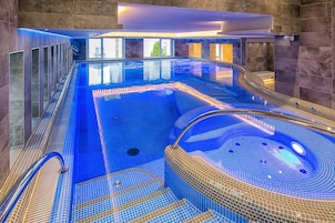 Uma grande piscina interior com uma banheira de hidromassagem, paredes azulejadas e vista clara para o exterior.