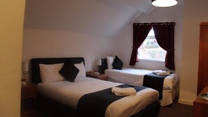 Quarto de hotel com duas camas, uma janela com cortinas, mesa de cabeceira com abajur e luminária no teto.