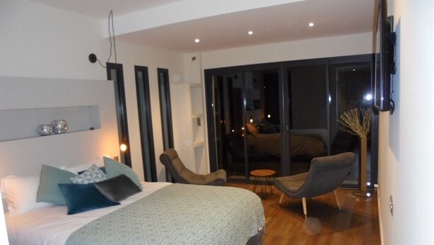 Quarto moderno com uma cama grande, duas poltronas, uma mesinha e varanda com vista para a cidade à noite.