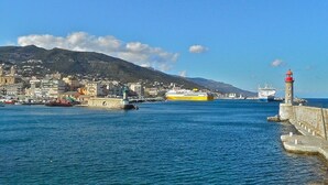 Uma cidade costeira com um farol, um grande navio amarelo e montanhas ao fundo.