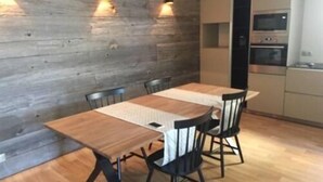 Uma sala de jantar com mesa e cadeiras de madeira, uma cozinha com eletrodomésticos embutidos e uma parede de pedra com iluminação embutida.
