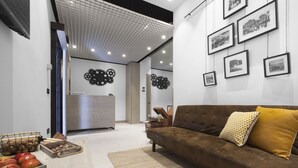 Uma sala de estar moderna com um sofá marrom, uma mesa de centro de madeira e um tapete. Há quadros nas paredes e um teto com padrão quadriculado.