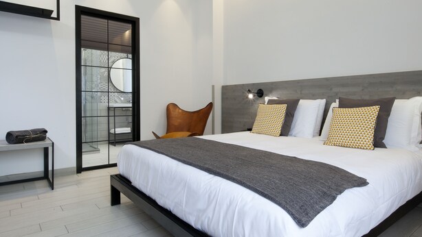 Quarto moderno com uma cama grande, mesa de cabeceira, uma cadeira e porta de vidro que dá para um banheiro.