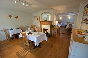 Uma sala de jantar com mesas e cadeiras de madeira, lareira e área de cozinha com uma fruteira.