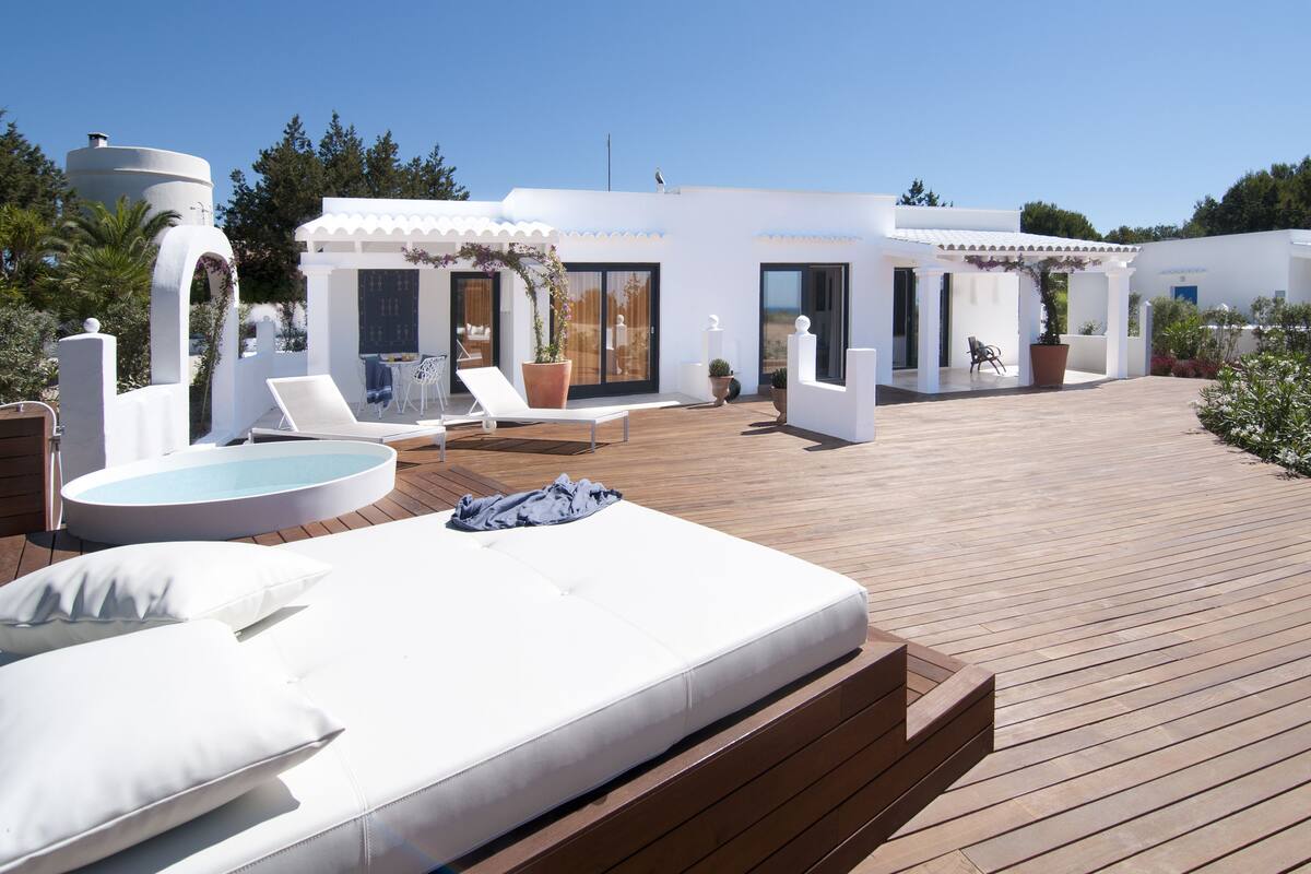 Uma área de estar ao ar livre moderna com um deck de madeira, cadeiras de lounge brancas, uma pequena piscina e vasos de plantas.