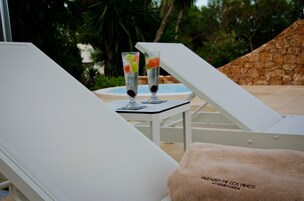 Uma área à beira da piscina com espreguiçadeiras e uma mesa com dois copos de um suco de fruta.