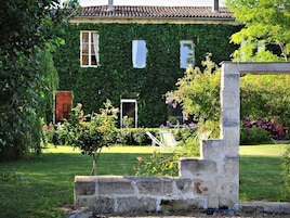 Uma casa com exterior coberto de hera verde, porta visível e jardim com gramado e algumas plantas.