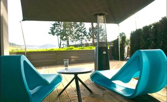 Área de estar coberta ao ar livre com duas cadeiras azuis e uma mesa pequena, com vista para um jardim paisagístico e colinas ao longe.