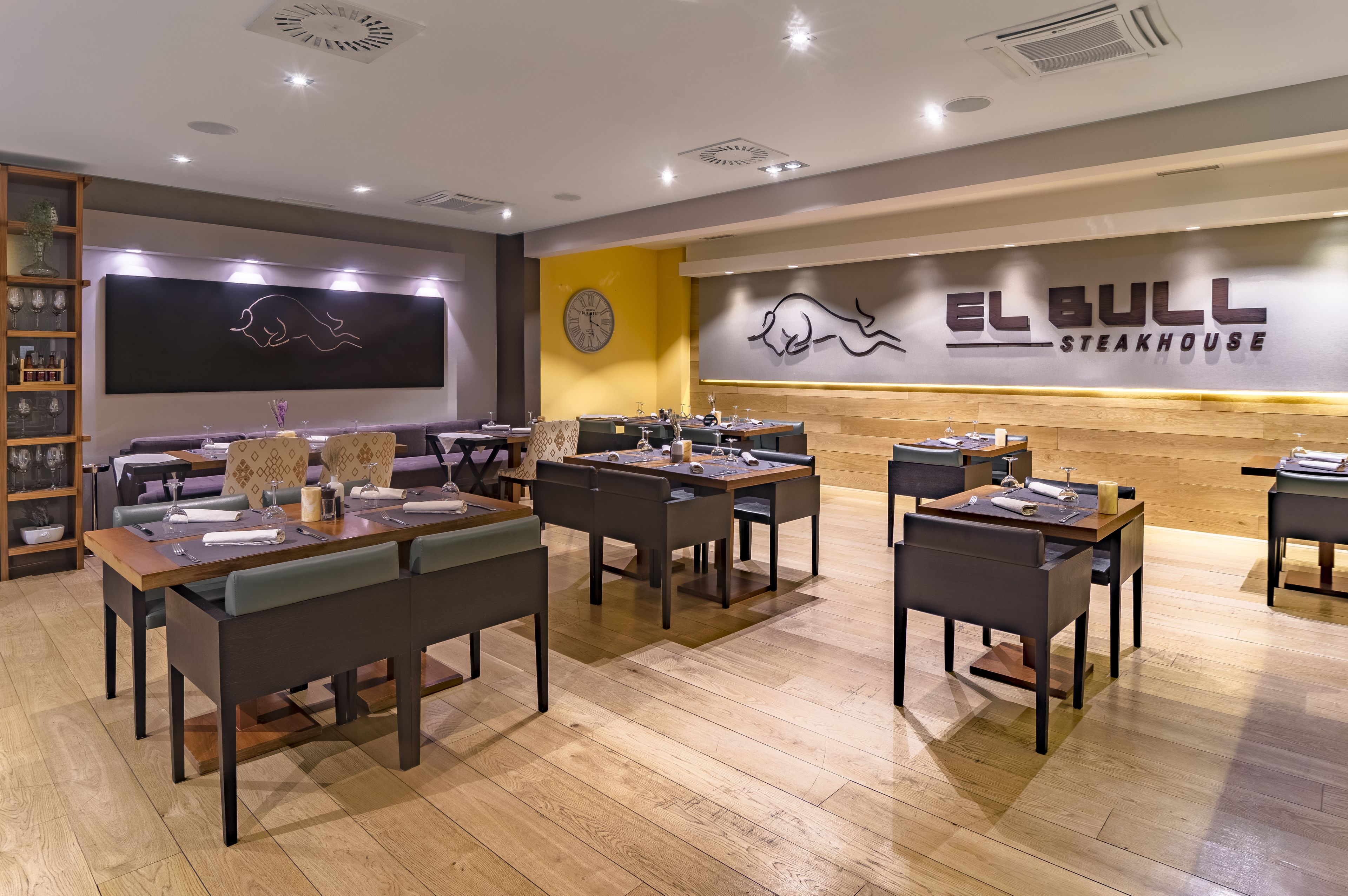 Interior de restaurante com mesas e cadeiras de madeira, um relógio e uma placa na parede que diz 'EL BULL STEAKHOUSE'.