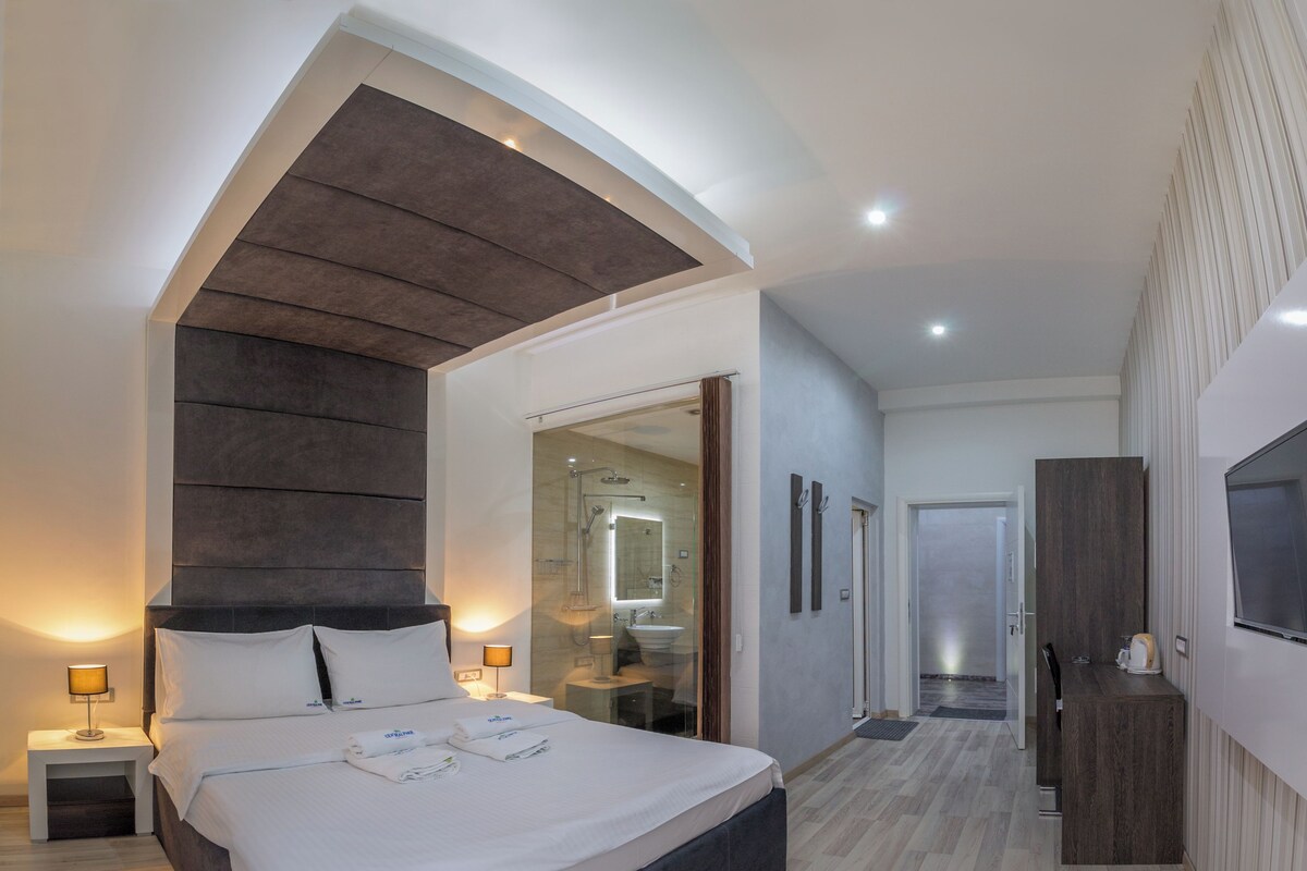 Quarto de hotel moderno com uma cama grande, mesa de cabeceira com luminária, box de vidro para o chuveiro, guarda-roupa e televisão fixada na parede.