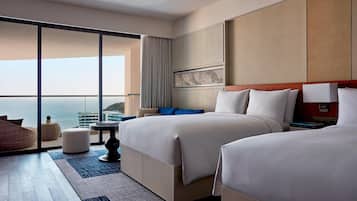 Quarto de hotel com duas camas, uma janela ampla com vista para o oceano, um espaço de estar com sofá e banquinho, e uma mesa pequena.