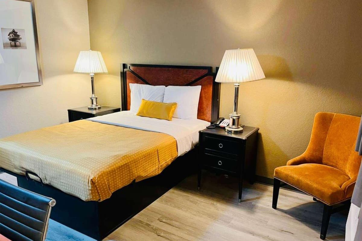 Quarto de hotel com cama, duas mesas de cabeceira com luminárias, uma cadeira e um quadro na parede.