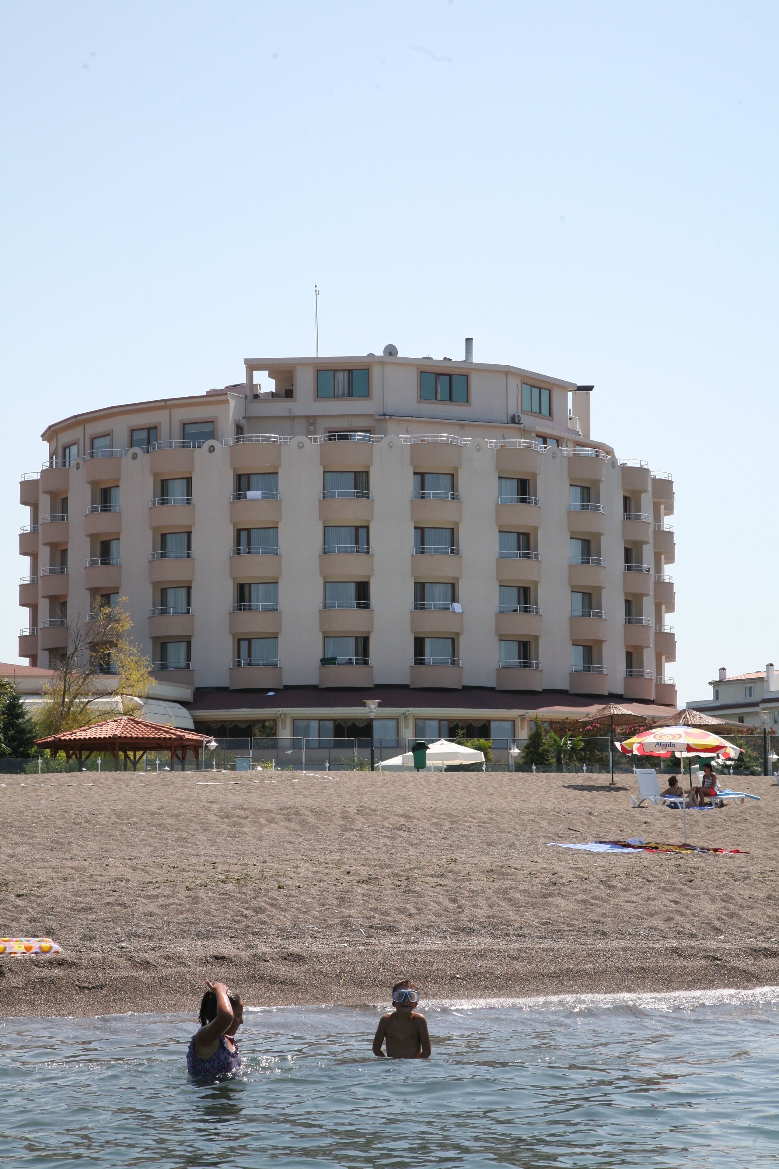 Um hotel de vários andares com varandas, uma praia em primeiro plano e um céu claro.