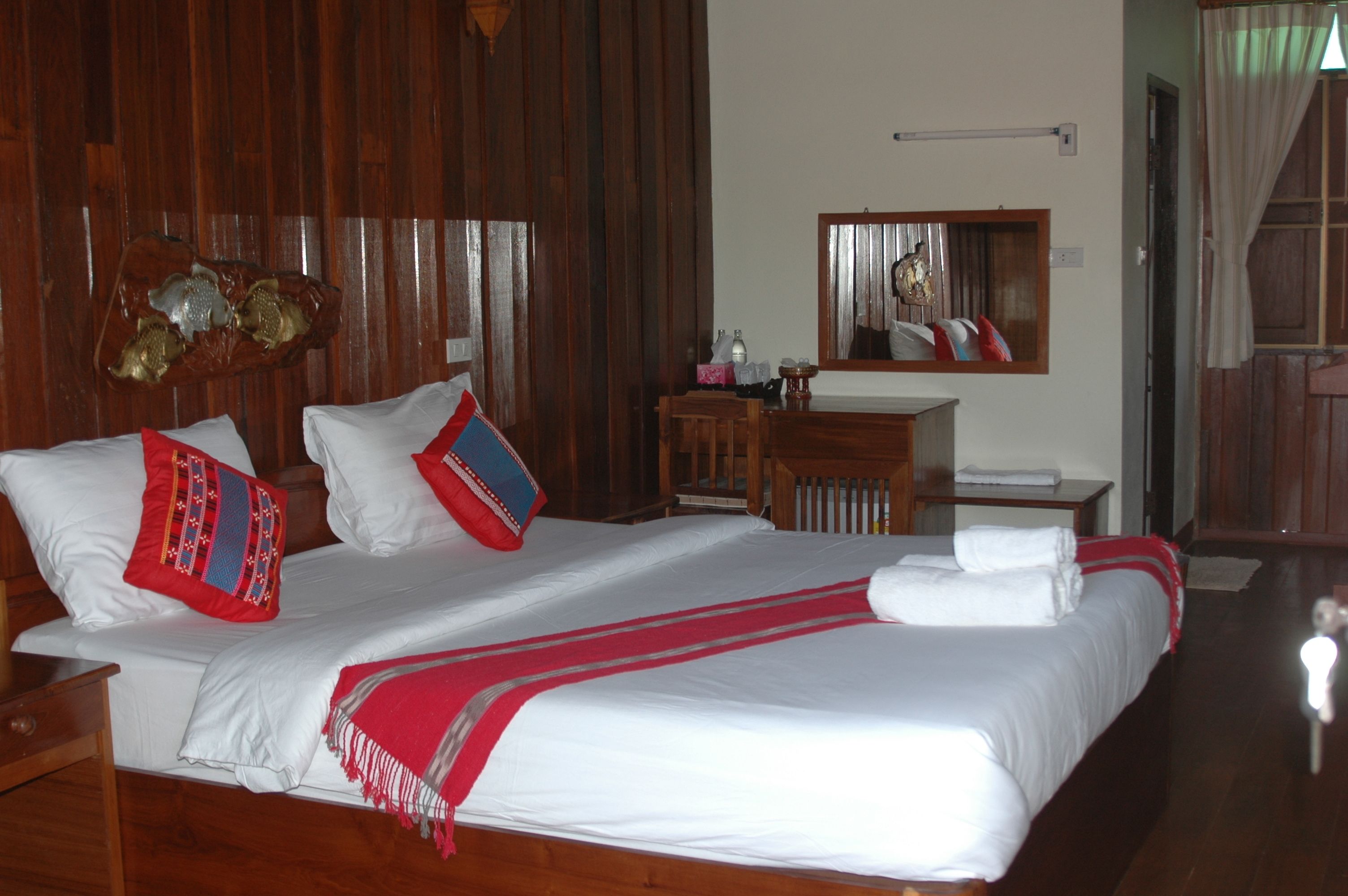 Cama bem arrumada com lençóis brancos e almofada vermelha e azul. Apresenta uma mesinha de cabeceira de madeira com um abajur e uma pequena mesa com um espelho em cima.