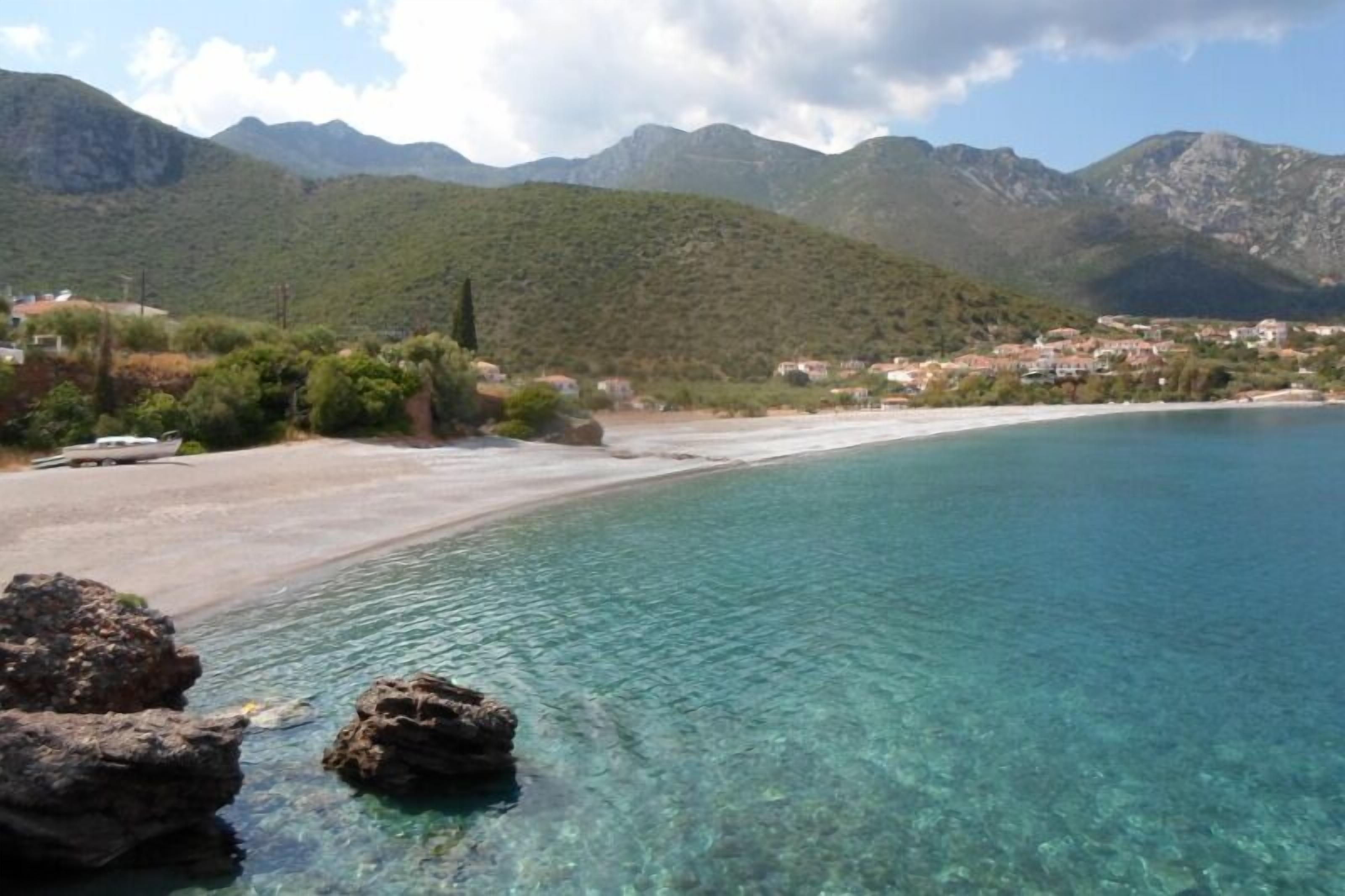 Paisagem costeira com águas turquesa cristalinas, afloramentos rochosos e um cenário montanhoso ao fundo.