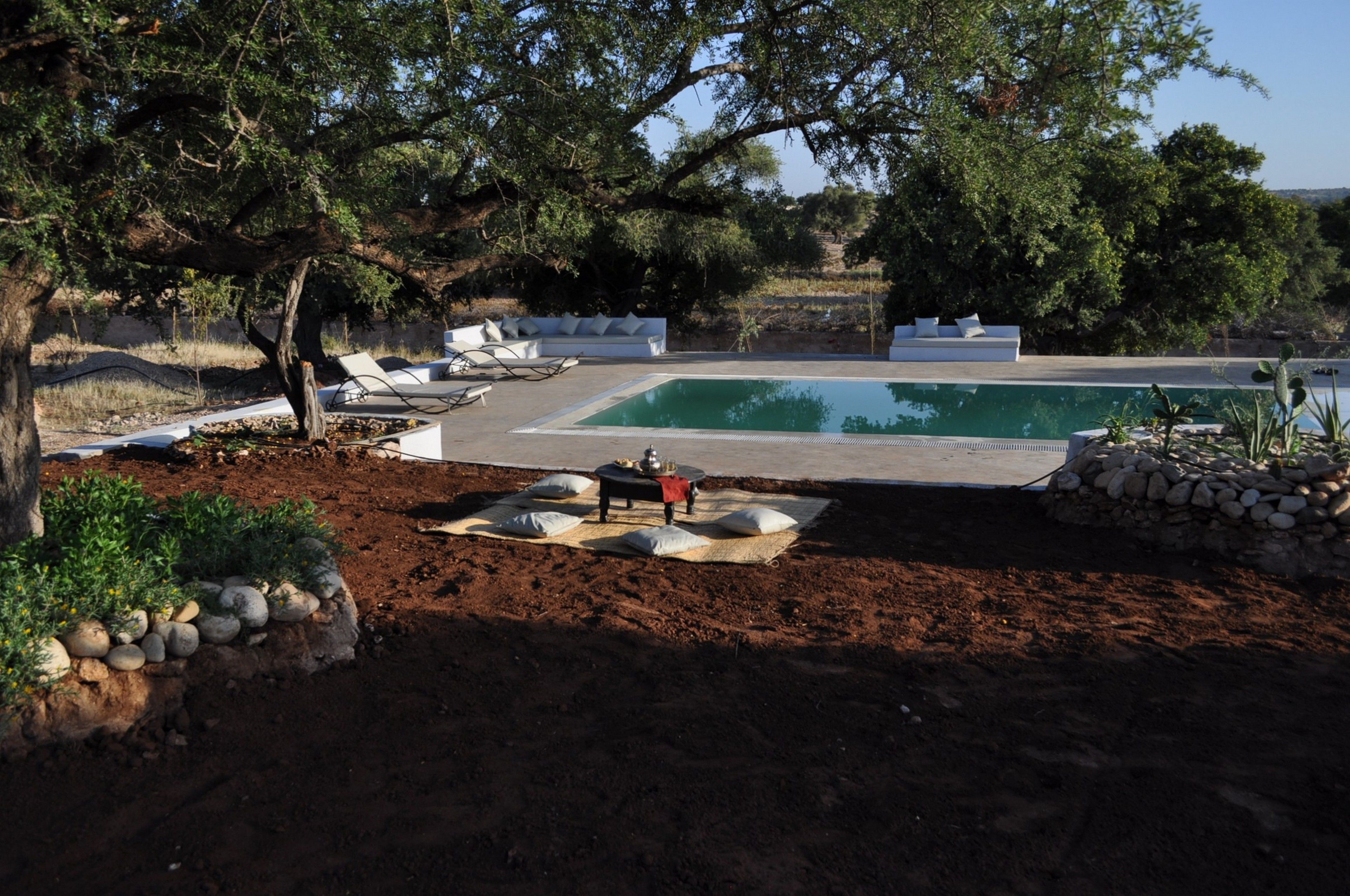 Uma área de piscina com espreguiçadeiras e uma mesa, cercada por árvores e chão de terra.