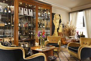 Quarto bem iluminado e elegante, com área de bar que apresenta um display de vinhos e copos, assentos confortáveis e uma grande pintura na parede.