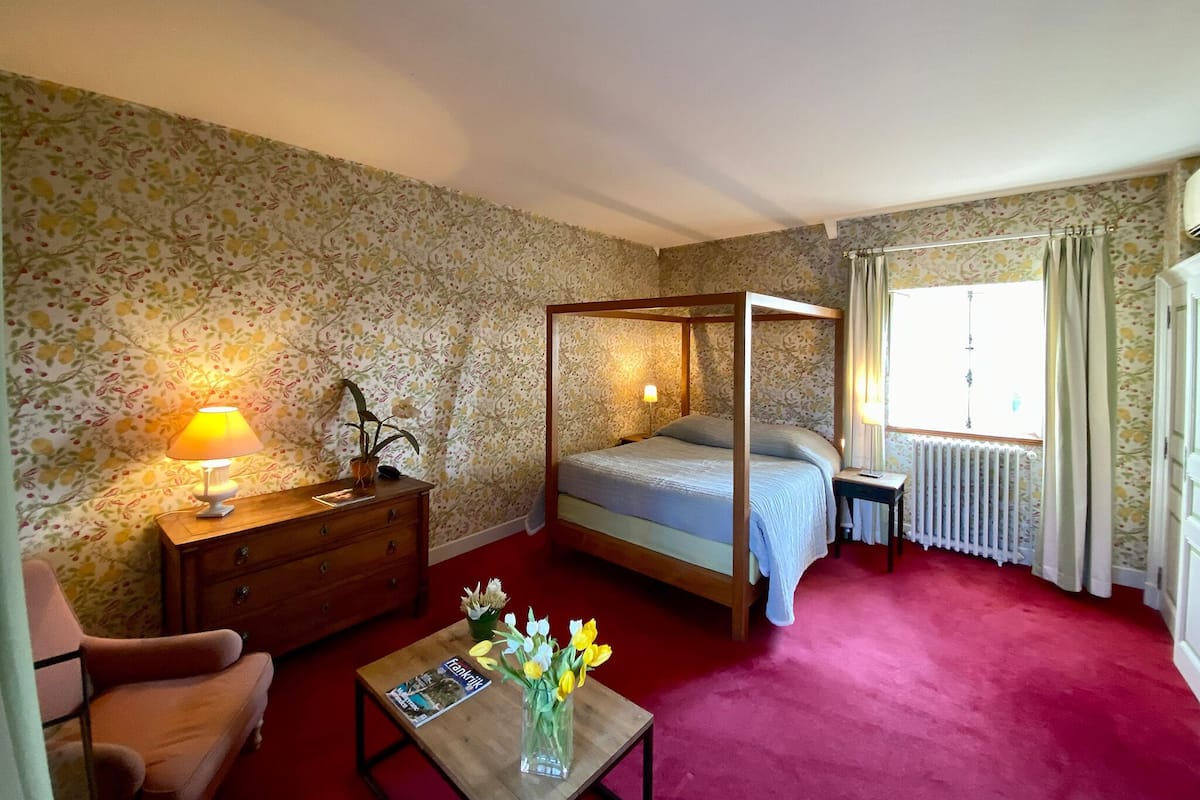 Quarto com cama de dossel, cómoda de madeira, cadeira, mesa com um vaso de flores e radiador.