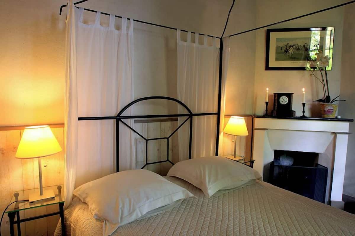 Quarto com cama, duas luminárias de cabeceira, um espelho, um relógio e uma lareira.