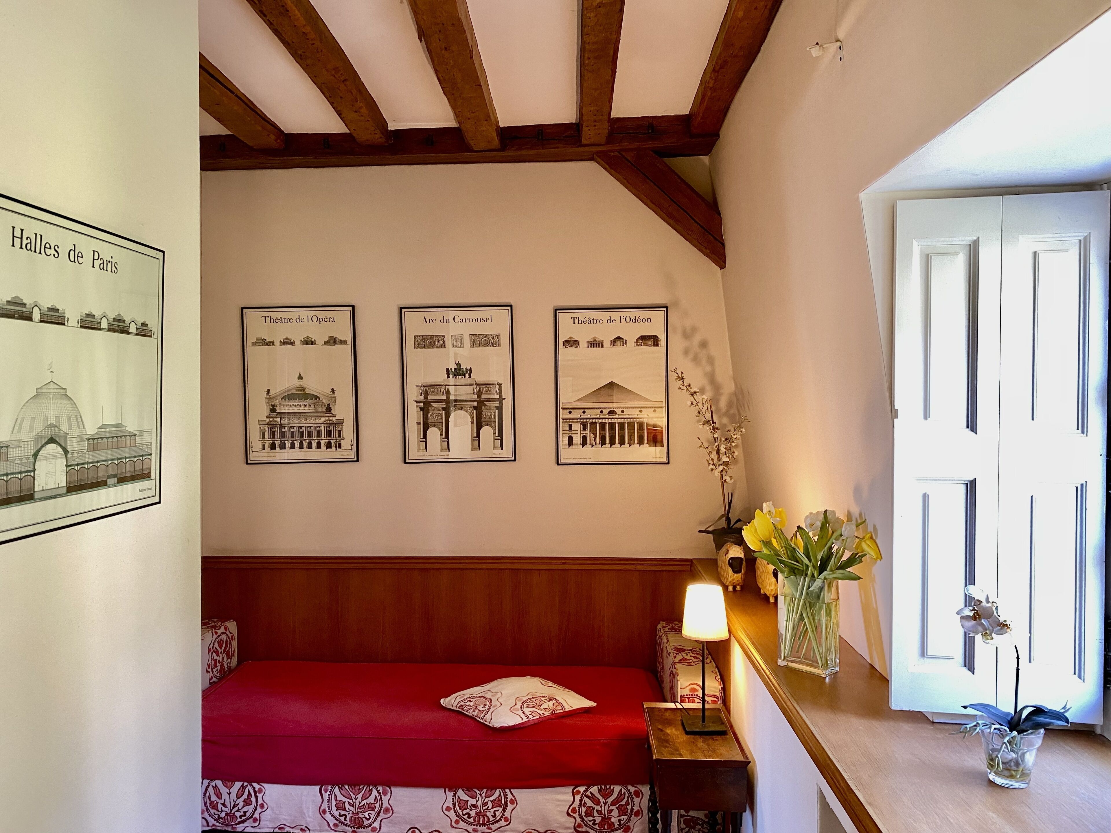 Quarto com cama vermelha, mesa de cabeceira com abajur e três quadros com imagens de pontos turísticos parisienses na parede.