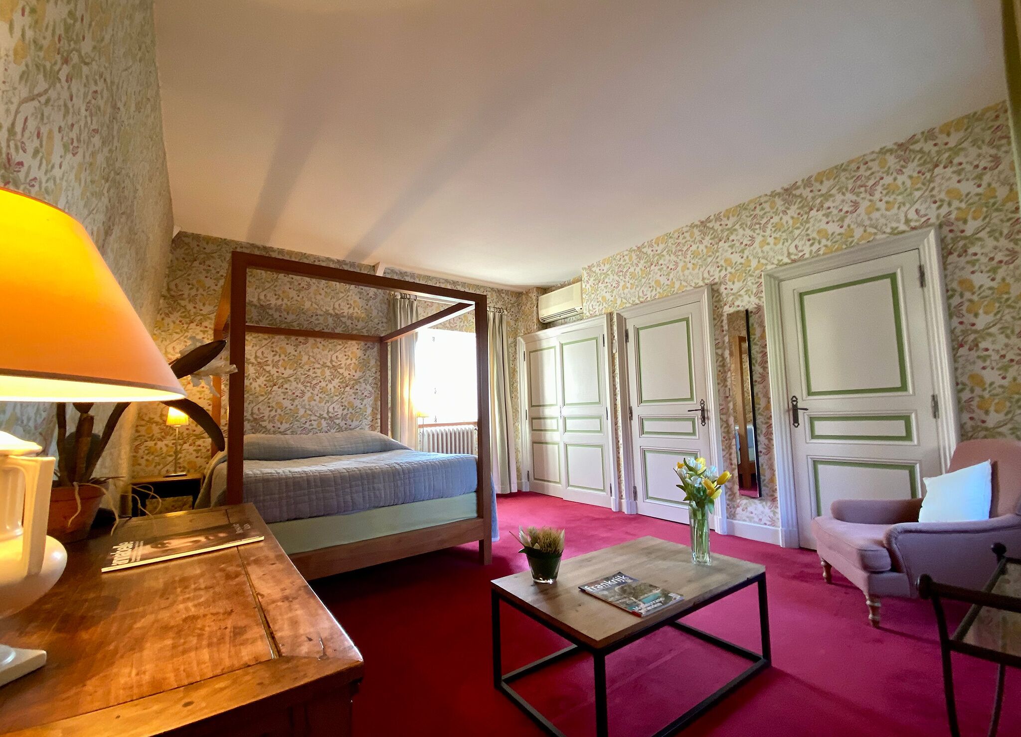 Quarto com cama de dossel, mesa de cabeceira de madeira, mesa com revista, cadeira e abajur.