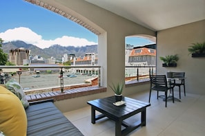 Varanda com vista para a paisagem urbana e montanhas. Há um sofá, uma mesa com uma planta e duas cadeiras.