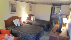 Quarto de hotel com duas camas, uma mesinha de centro com abajur, uma cadeira, uma escrivaninha com computador e um espelho.