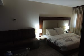 Quarto de hotel com cama, mesa de cabeceira, luminária, sofá e janela com cortinas.