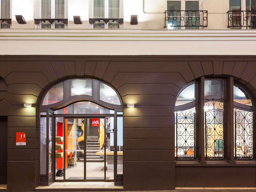 Entrada de um hotel com porta de vidro, escada que leva ao interior e uma varanda acima.
