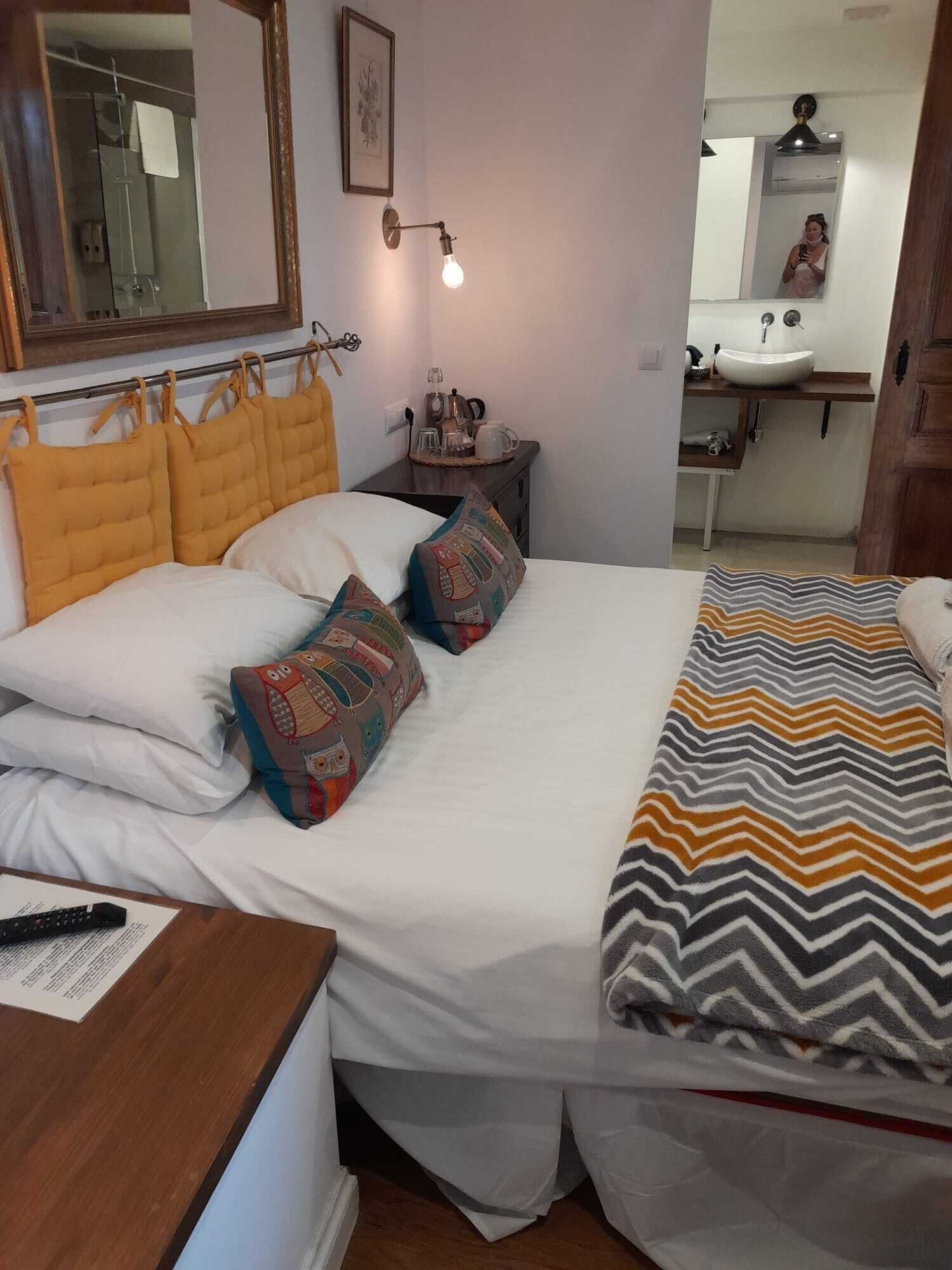 Quarto de hotel com uma cama, mesa de cabeceira com controle remoto, espelho, pequena mesa com cafeteira e banheiro visível por uma porta aberta.