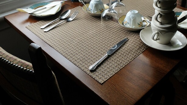 Uma mesa de jantar posta com jogo americano, pratos, talheres, um vaso com flores e um menu.