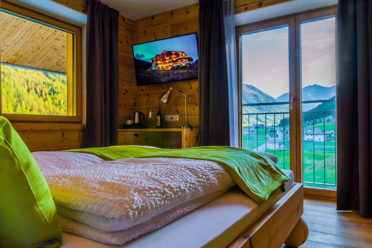 Quarto com uma cama, televisão fixada na parede e vista para uma paisagem montanhosa através de uma janela grande.