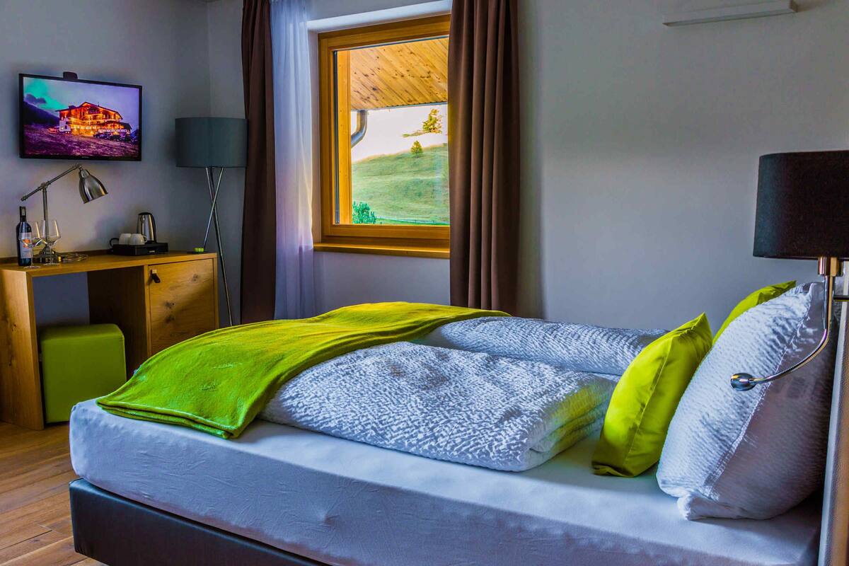 Quarto com cama, escrivaninha, televisão e janela com vista para uma casa.