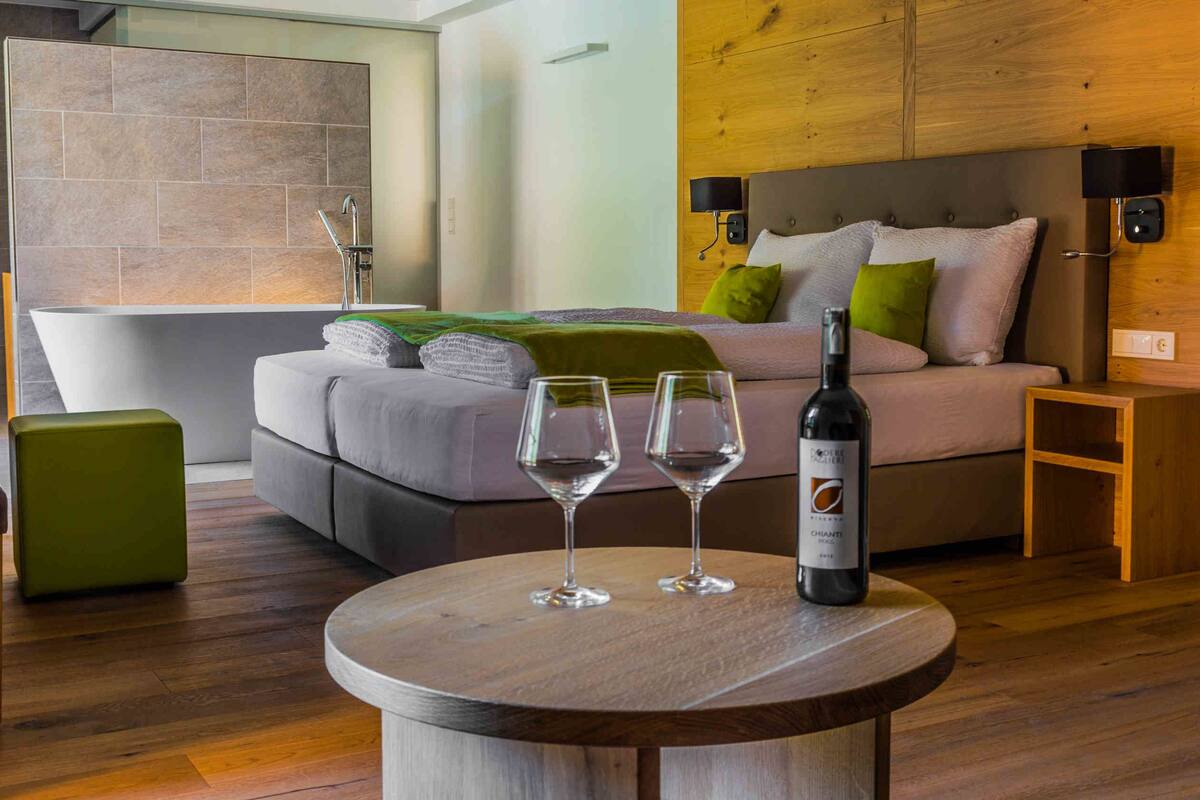 Quarto moderno com cama, mesas de cabeceira, uma mesa redonda de madeira com copos de vinho e uma garrafa de vinho.