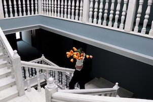 Uma escadaria imponente com balaústres brancos e um vaso de flores laranjas.