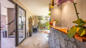 Um hall de hotel moderno com uma receção de pedra, área de estar com sofá e mesa de centro, uma estante de livros e vista para o jardim exterior.