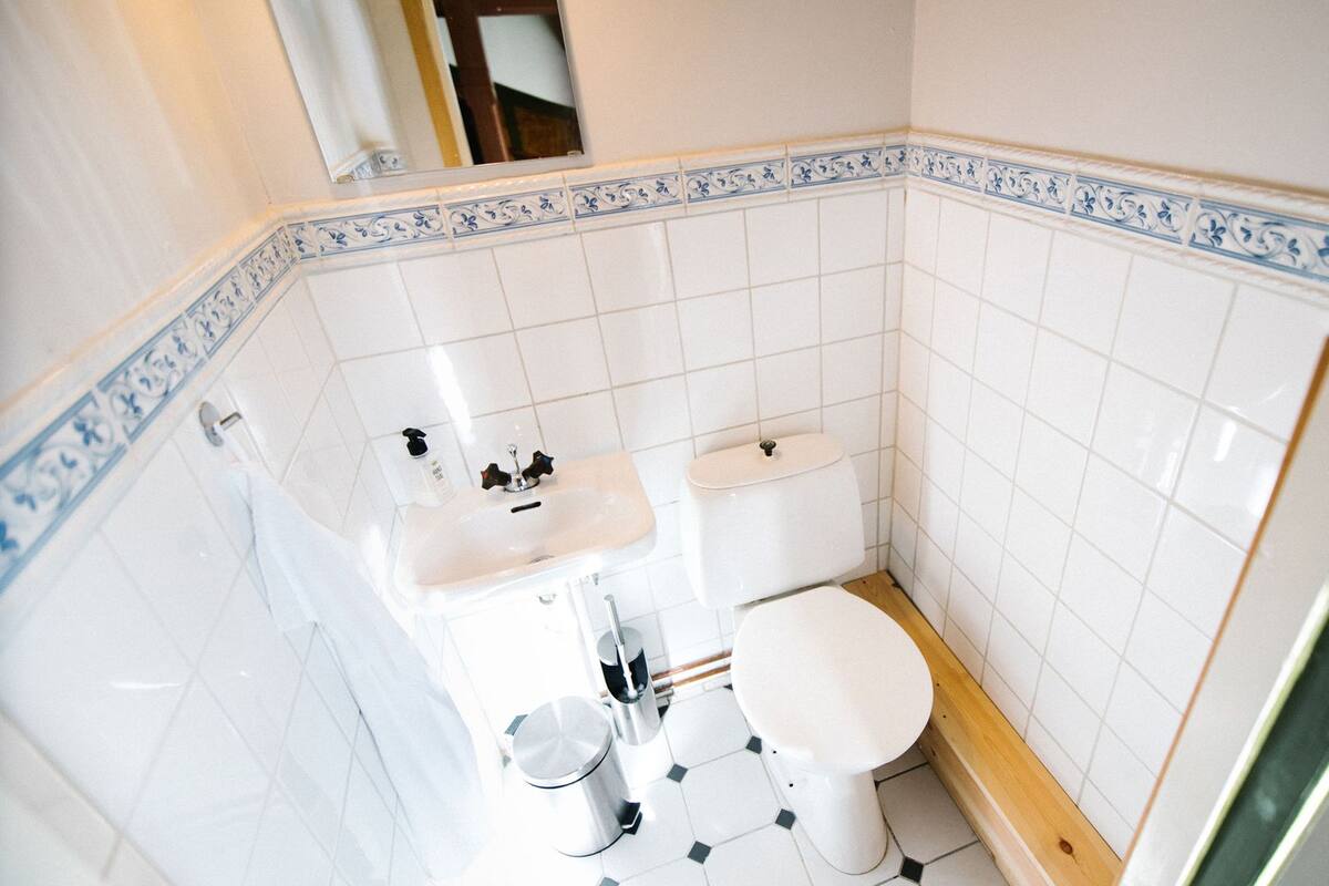 Uma casa de banho pequena com lavatório, sanita e bidé brancos, revestida a azulejo branco com detalhes decorativos azuis.