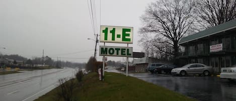 Placa de motel com o texto '11-E' e 'MOTEL' em verde, ao lado de uma estrada com carros estacionados e um edifício ao fundo.