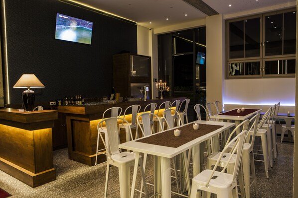 Restaurante com cadeiras brancas, área de bar e uma TV exibindo um evento esportivo.