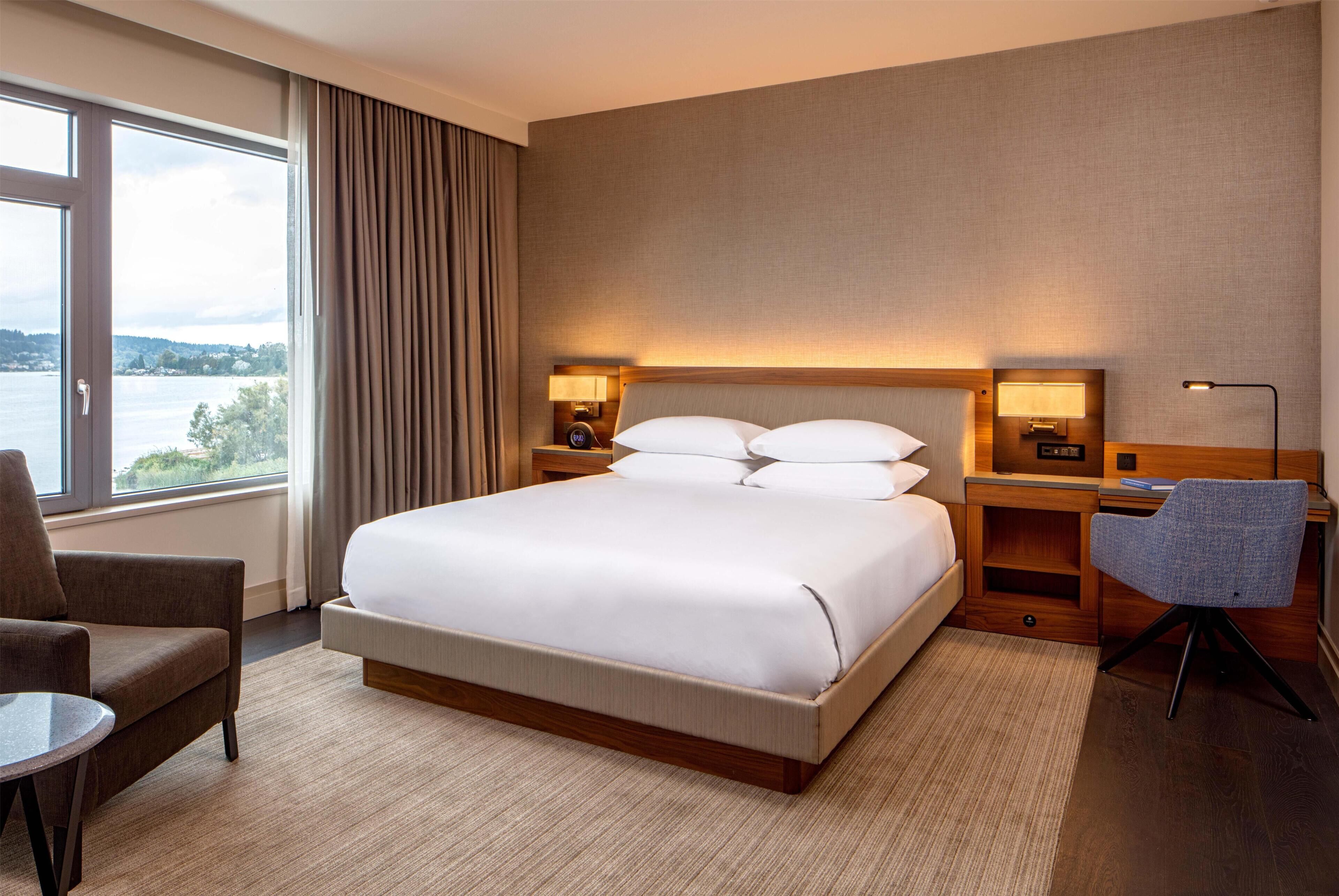 Quarto de hotel com uma cama grande, duas mesas de cabeceira com luminárias, uma cadeira e vista para um corpo de água.